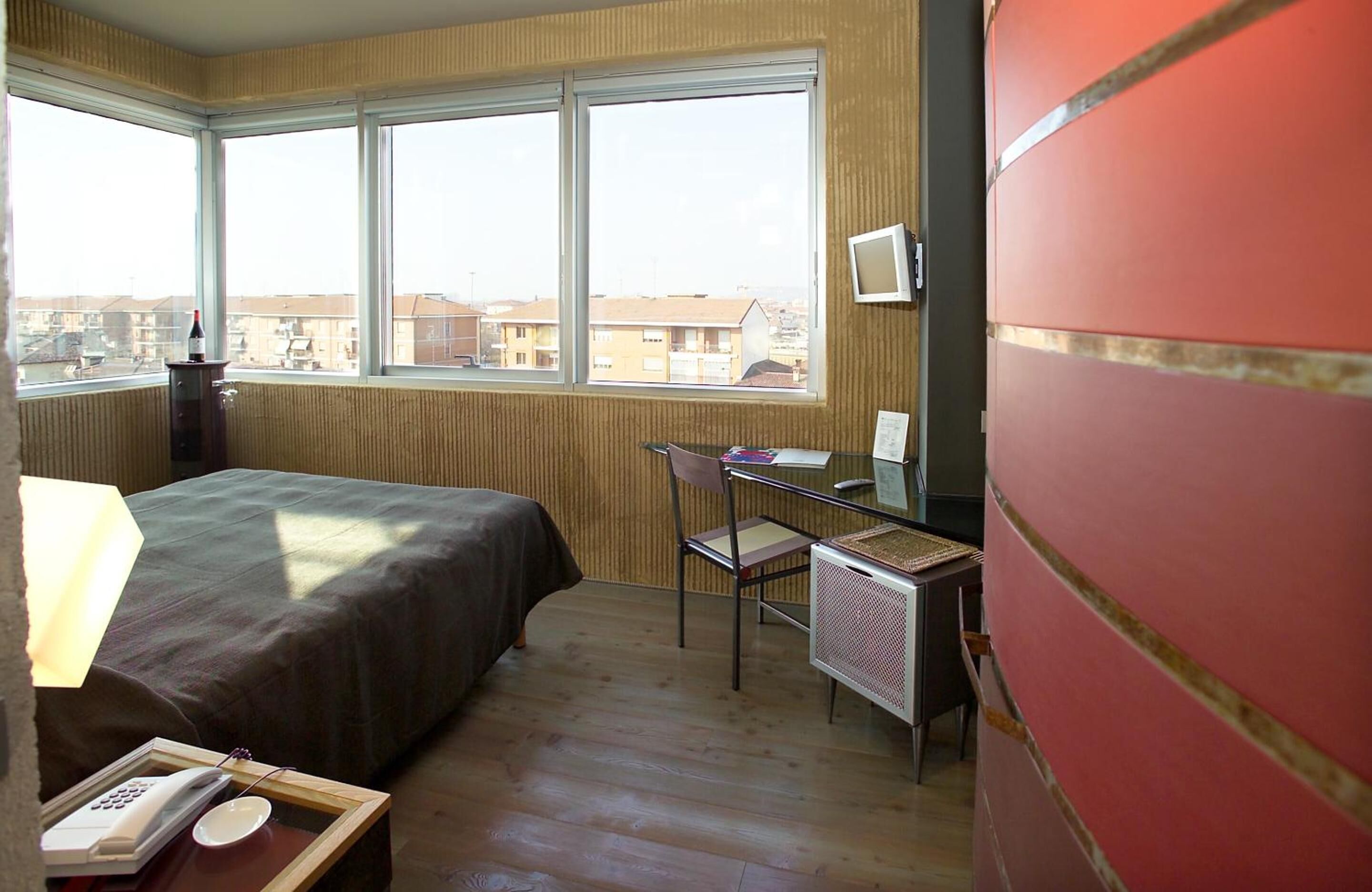 Quarto de hotel com cama, escrivaninha, cadeira, telefone e janela com vista para edifícios.
