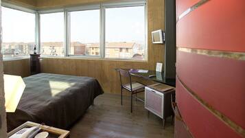 Quarto de hotel com cama, escrivaninha, cadeira, telefone e janela com vista para edifícios.