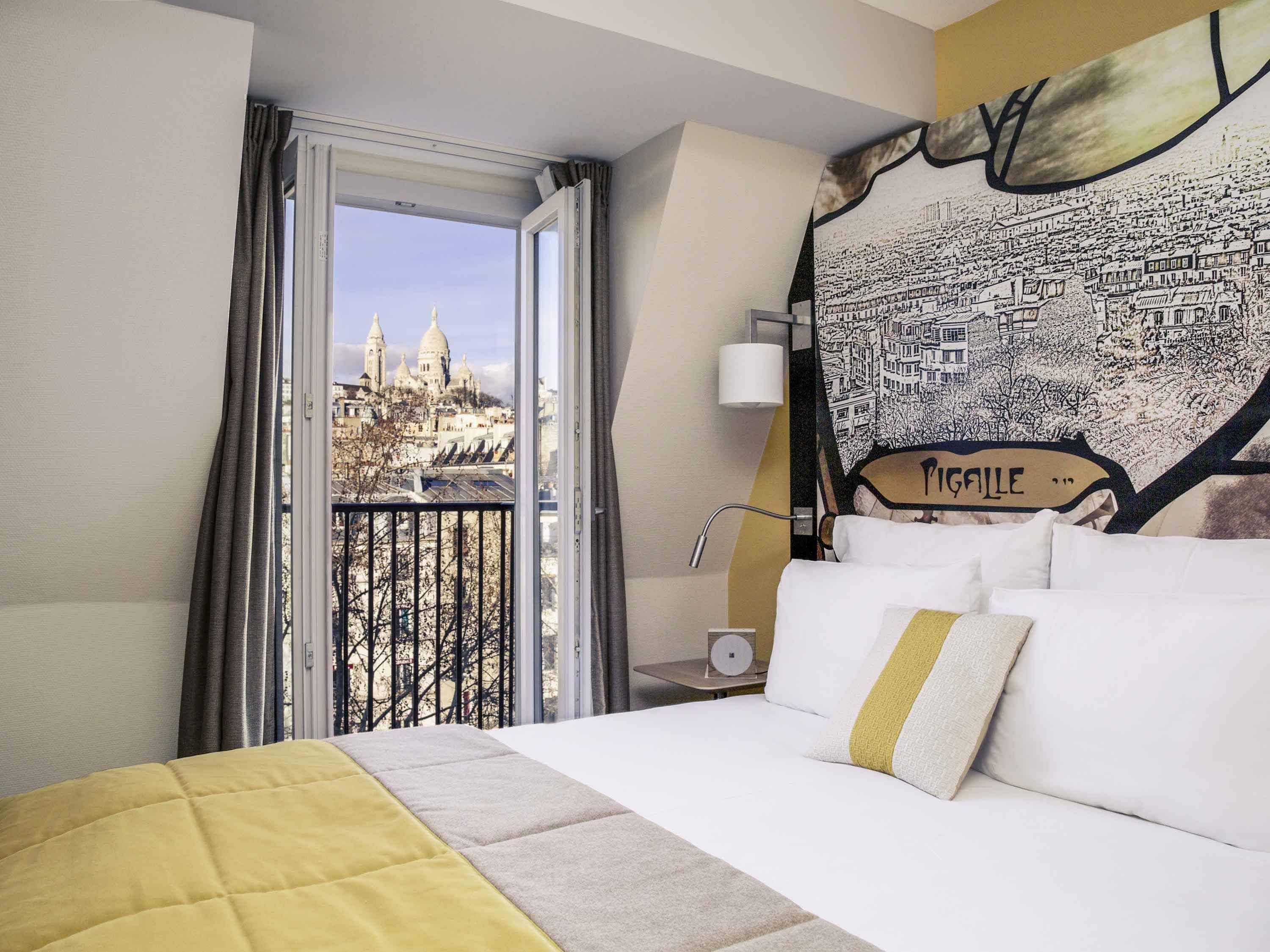 Quarto de hotel com cama, janela com cortinas e vista para o horizonte urbano, além de uma parede com um mural da paisagem da cidade.