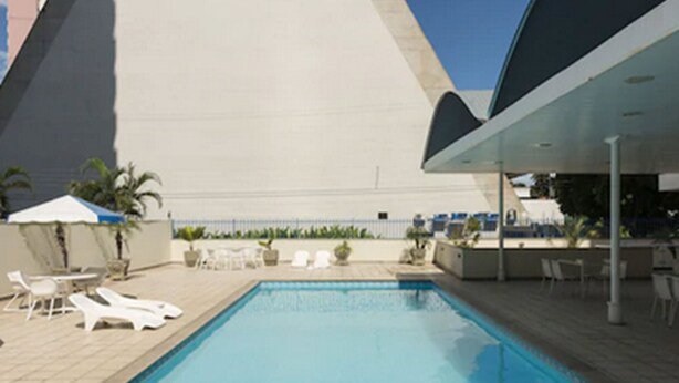Piscina com espreguiçadeiras e guarda-sóis, cercada por um deck ladrilhado e plantas em vasos.