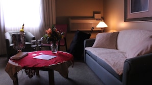 Quarto de hotel com toalha de mesa vermelha, sofá, poltrona, luminária e um vaso com flores.