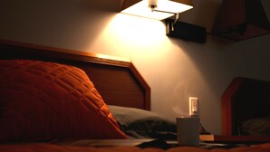 Quarto de hotel com iluminação fraca, uma cama, abajur e mesa de cabeceira com uma xícara.