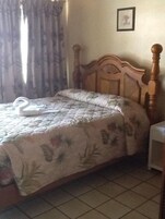 Quarto com estrutura de cama de madeira, roupa de cama florida, mesa de cabeceira branca e um quadro na parede.