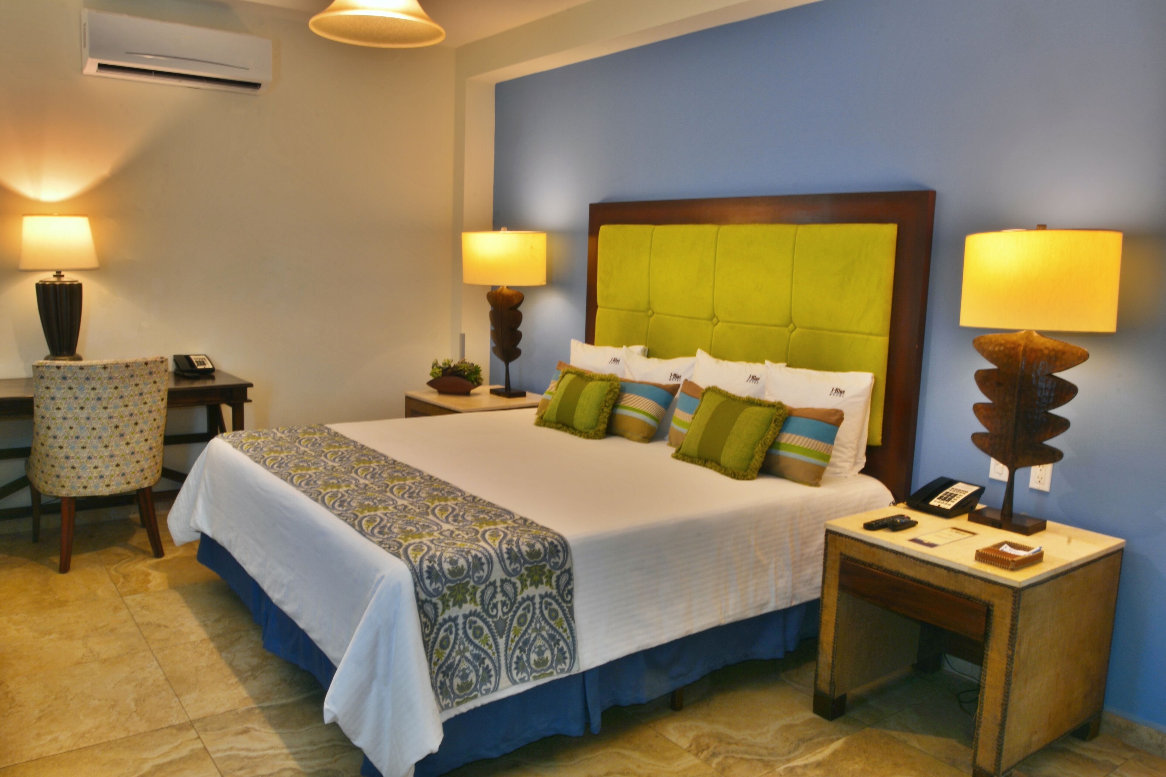 Quarto de hotel com uma cama grande, duas mesas de cabeceira com luminárias, uma cadeira e uma escrivaninha com telefone.