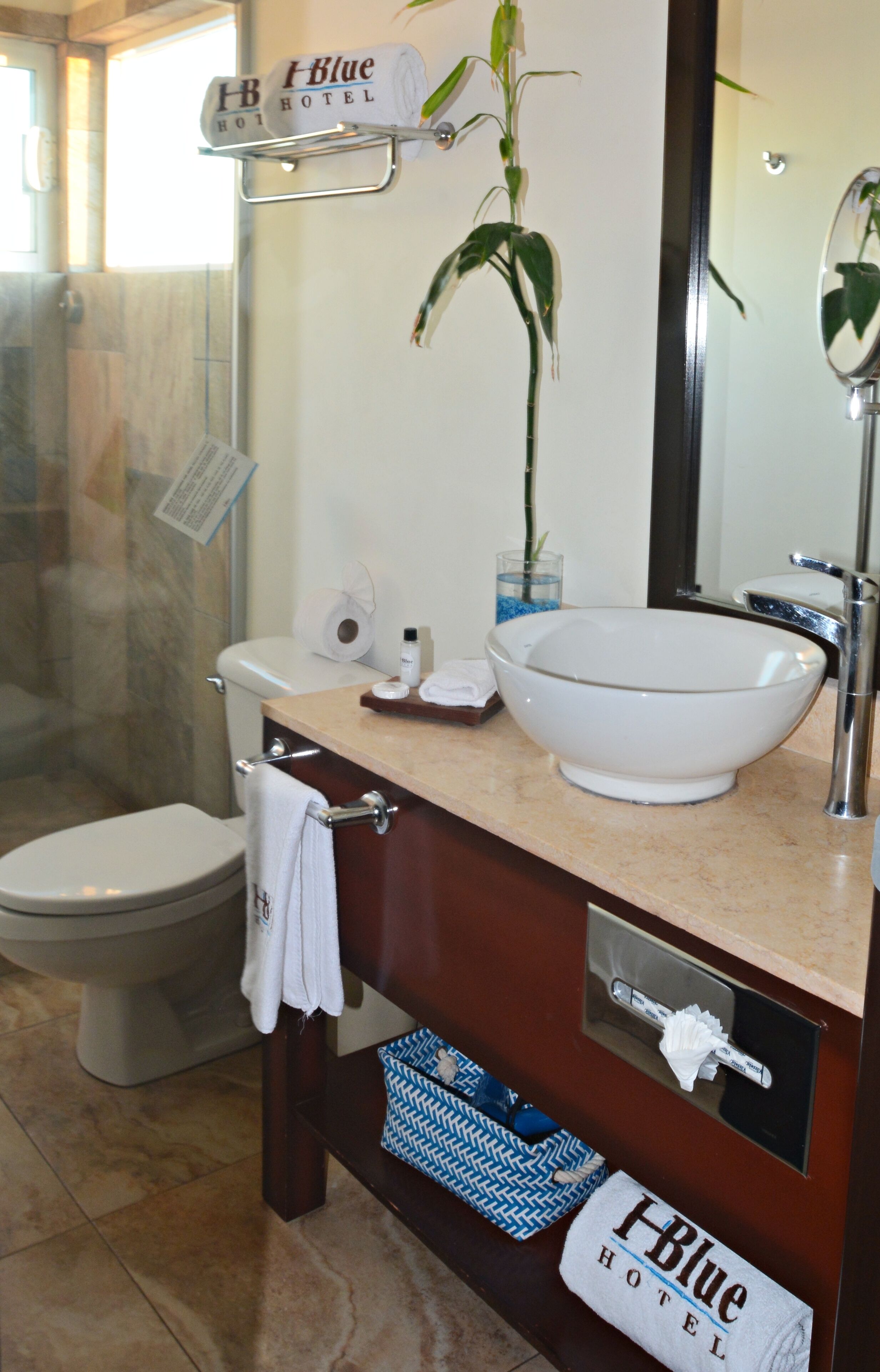 Casa de banho com sanita, lavatório, espelho e chuveiro. Há um porta-toalhas com uma toalha e uma planta.