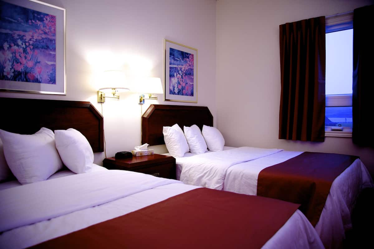 Quarto de hotel com duas camas, uma mesa de cabeceira, uma luminária e duas imagens emolduradas na parede.