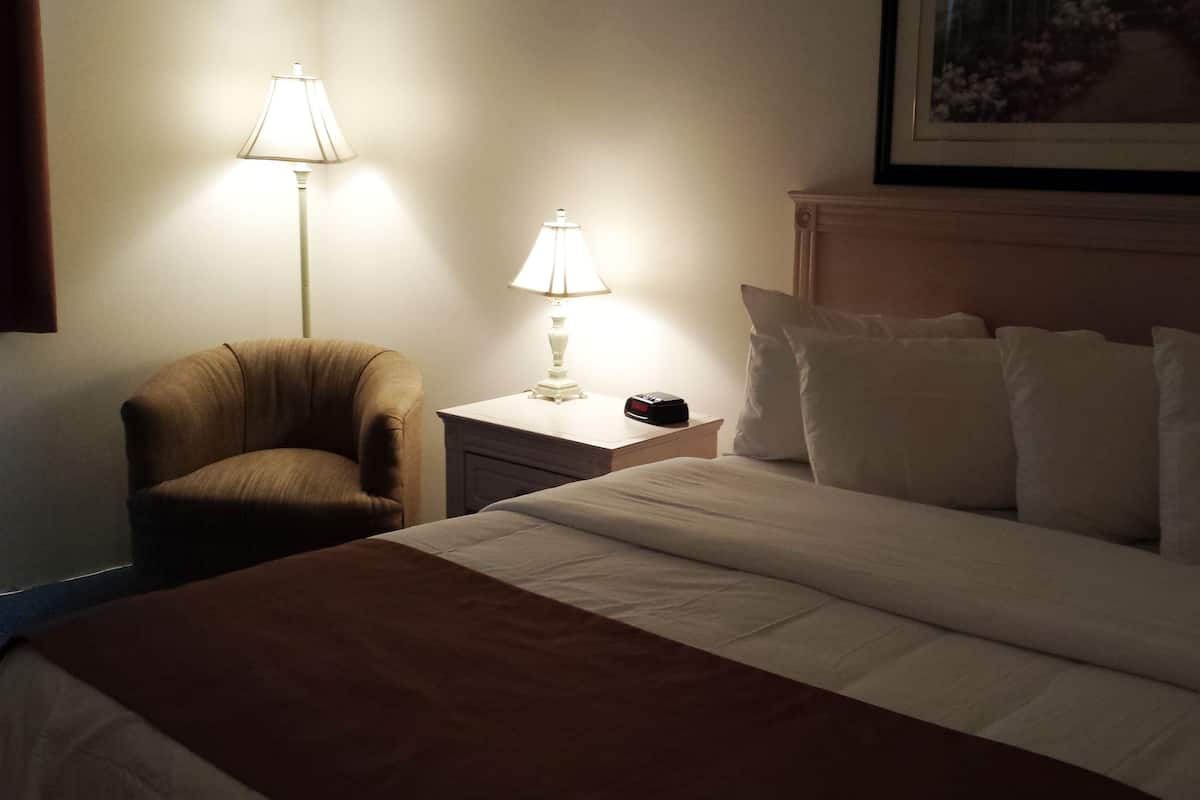 Quarto de hotel com uma cama, duas luminárias de cabeceira, uma cadeira, mesa de cabeceira com relógio e um quadro na parede.
