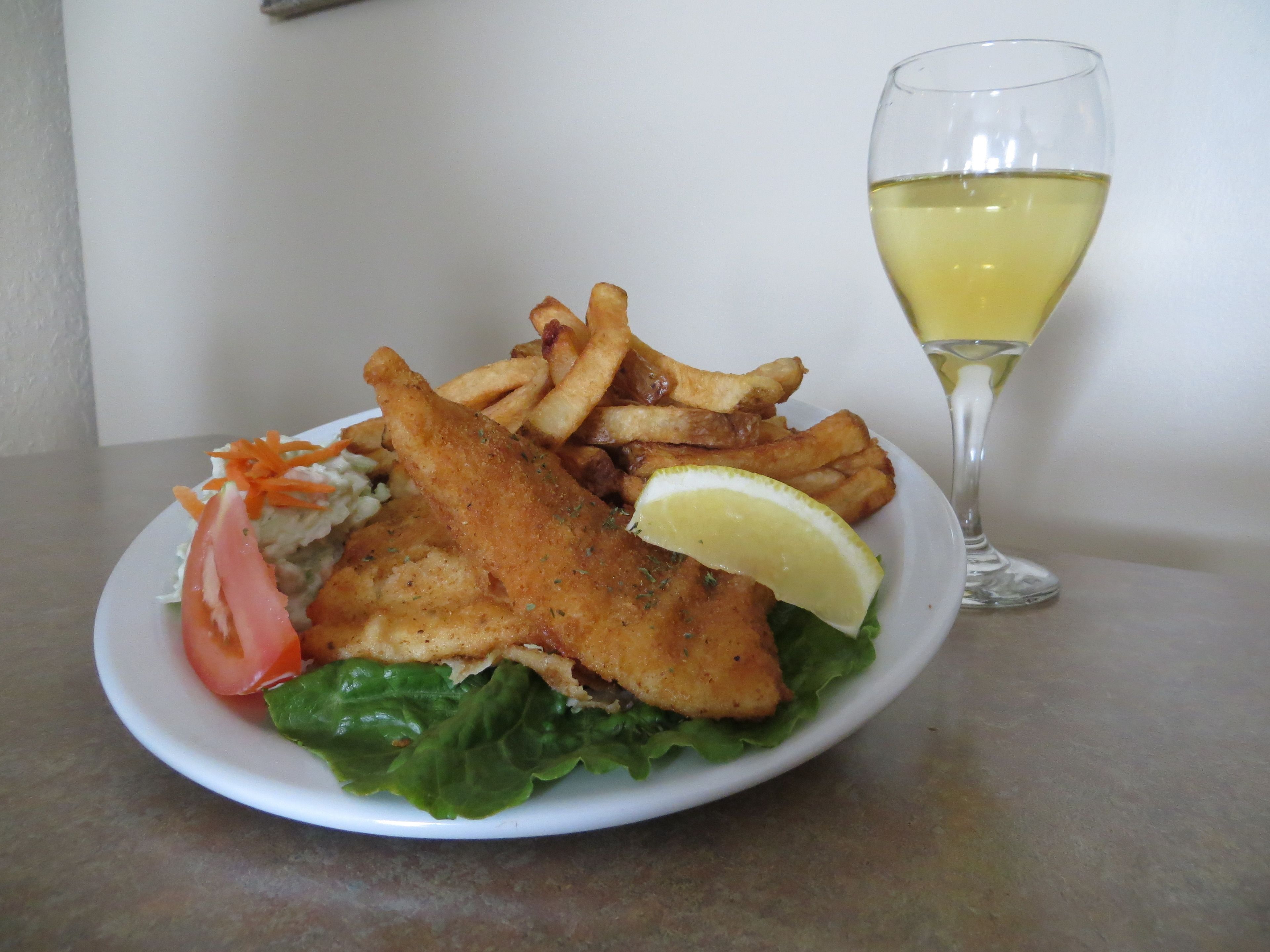 Um prato de peixe com batatas fritas, uma salada, uma fatia de limão e um copo de vinho branco.