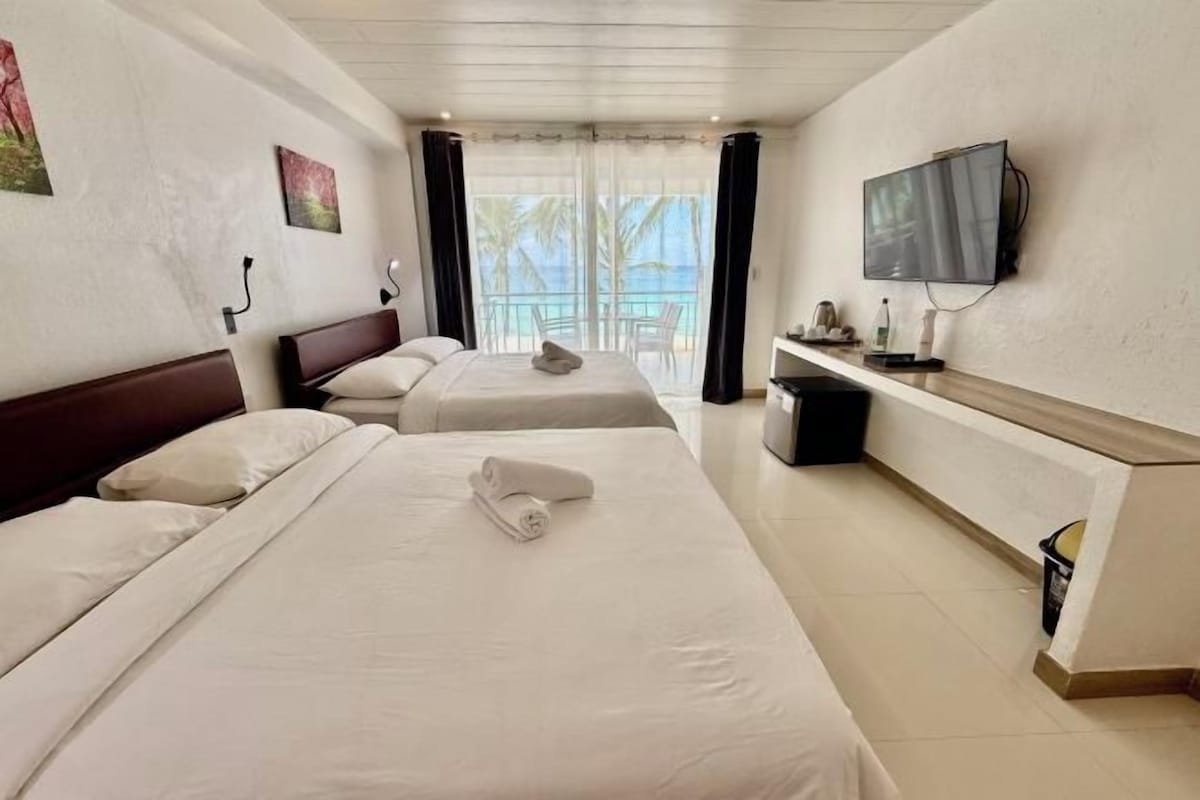 Quarto de hotel com duas camas, televisão, varanda com vista para o mar e uma pequena mesa de trabalho com cadeira.