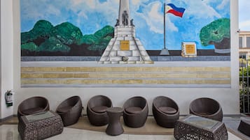 Um mural retrata um monumento com inscrições, uma bandeira e uma estátua. Em frente, há cadeiras de vime e uma mesa de vidro.