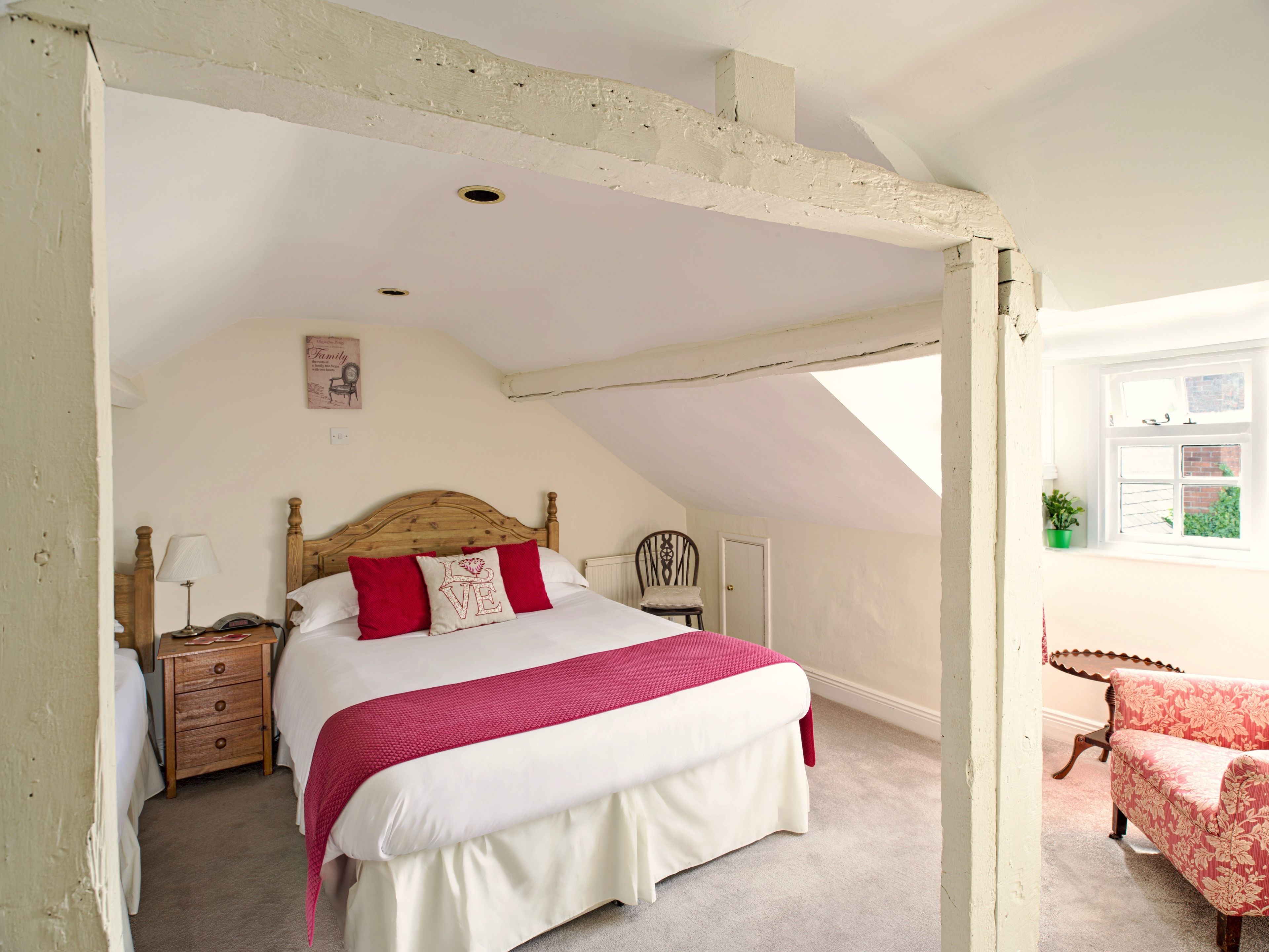 Quarto com cama de madeira, roupa de cama vermelha e branca, uma mesinha de cabeceira e uma cadeira.