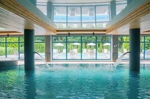 Piscina interior com água cristalina, rodeada por grandes janelas e espreguiçadeiras brancas.