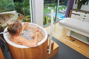 Uma banheira de hidromassagem cheia de água e pétalas de rosa, piso de madeira e vista para uma piscina e jardim através de grandes janelas.