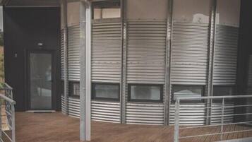 Um edifício com deck de madeira, persianas de metal e rampa de concreto, além de corrimão metálico.