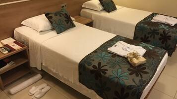 Quarto de hotel com duas camas, cada uma com colcha estampada com flores, uma mesinha de cabeceira com controle remoto e um par de chinelos no chão.