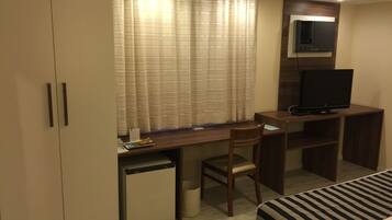 Quarto de hotel com escrivaninha, televisão, pequeno frigorífico, cadeira e cama com roupa de cama listrada.