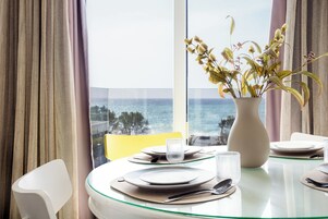 Uma mesa de jantar com pratos, talheres e um vaso de flores, em frente a uma janela com vista para o mar.