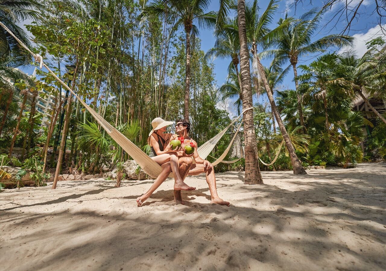 Um casal sentado numa rede em um cenário tropical, com palmeiras e uma praia arenosa.