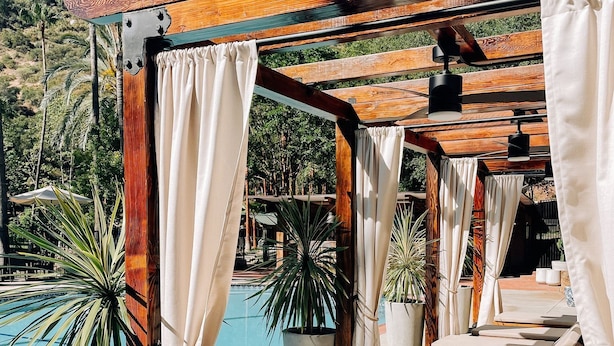 Uma pérgola de madeira com cortinas brancas, uma área de estar junto à piscina e plantas em vasos.