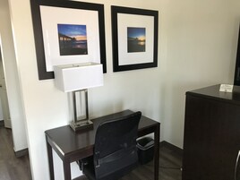 Quarto de hotel com escrivaninha, uma cadeira, abajur, lixeira e duas quadros na parede.