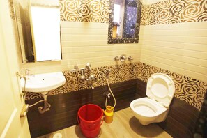 Casa de banho com lavatório, sanita e chuveiro. Há um balde vermelho e um recipiente amarelo no chão.