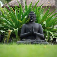 Estátua de uma figura sentada em pose meditativa, rodeada por folhagem verde exuberante.