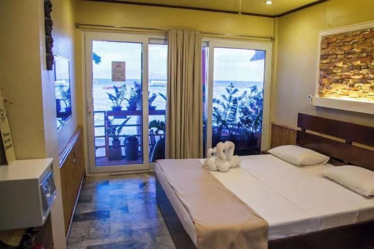 Quarto de hotel com cama, escrivaninha, cadeira e vista para o mar através de uma porta deslizante.