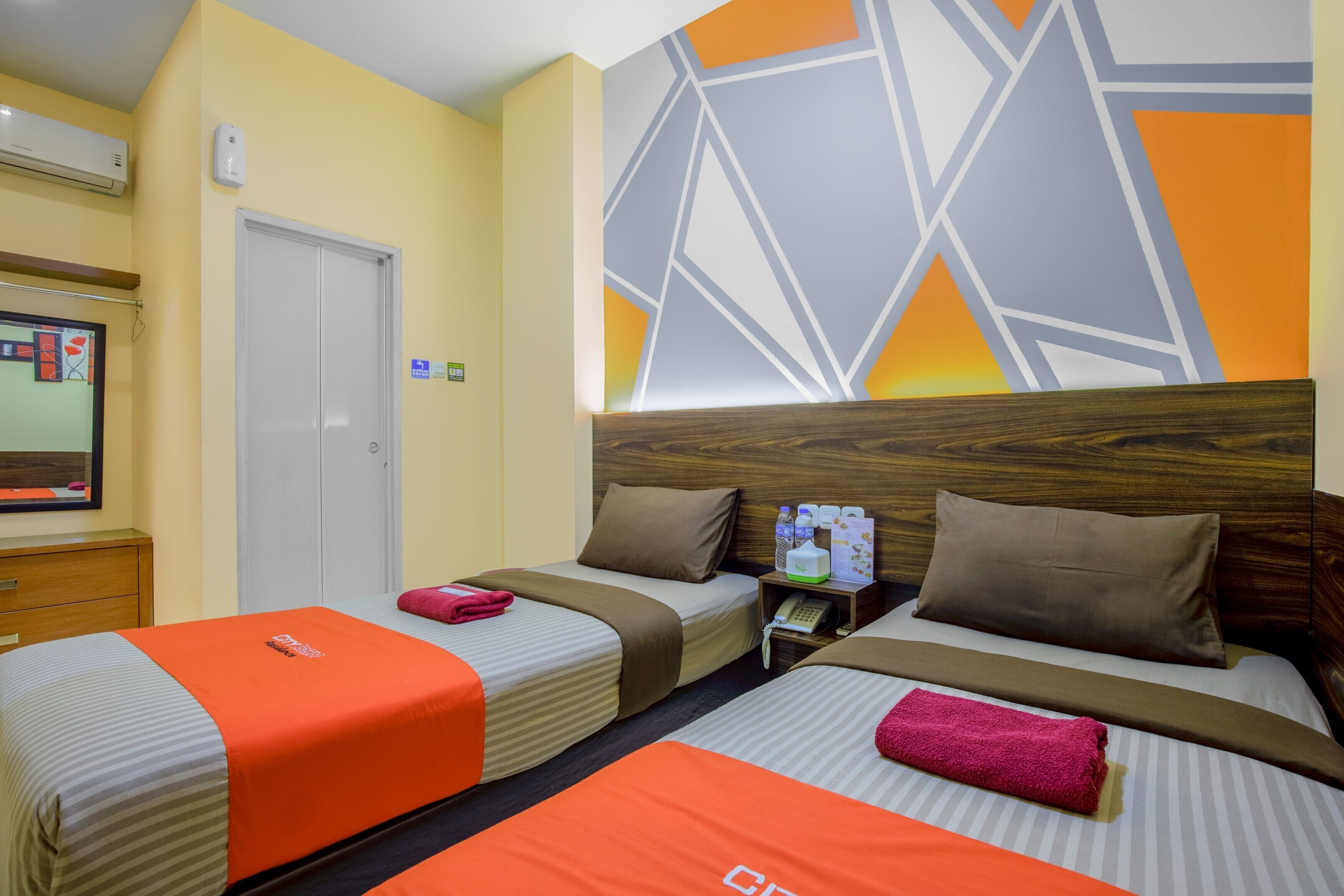 Quarto de hotel com duas camas, cada uma com colcha listrada e detalhes em laranja. Há uma cabeceira de madeira, uma mesa de cabeceira com telefone e artigos de higiene pessoal, e uma parede com padrões geométricos.