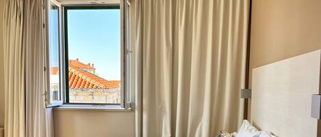 Quarto de hotel com cama, janela com cortinas, vista para edifícios e um vaso com flores na mesa de cabeceira.