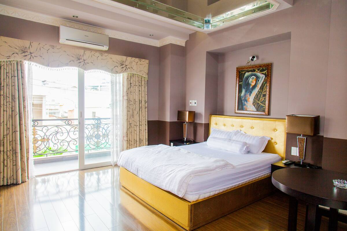 Quarto de hotel com uma cama grande, varanda com cortinas, um quadro na parede e uma mesinha com abajur.