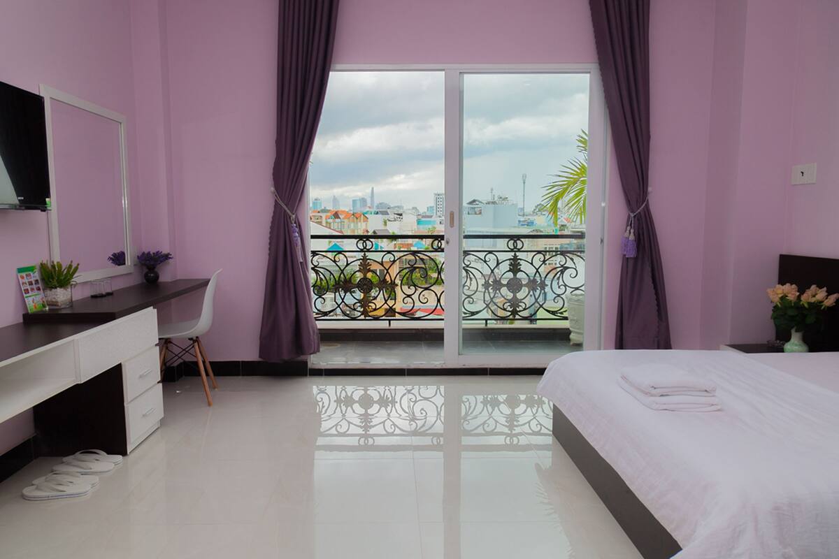 Quarto com cama, escrivaninha, cadeira, cômoda e varanda com vista para a cidade.