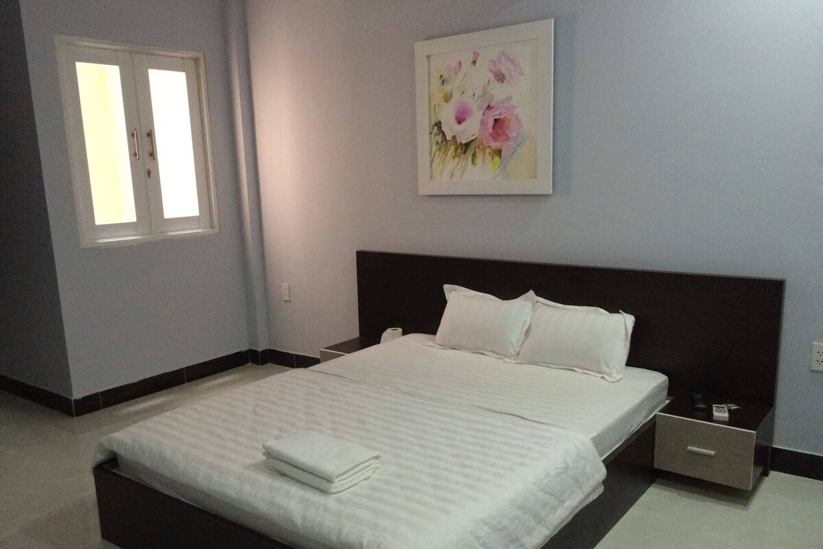 Quarto com cama, mesa de cabeceira, gaveta, janela e um quadro na parede.