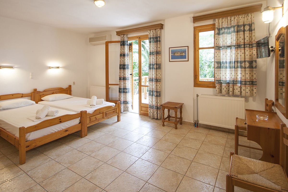 Quarto de hotel com duas camas de madeira, uma mesa pequena de madeira, uma cadeira e uma janela com cortinas.