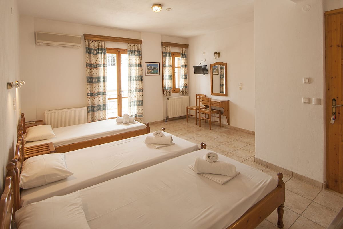 Quarto de hotel com duas camas de solteiro, uma escrivaninha de madeira, uma cadeira, um radiador e uma televisão na parede.