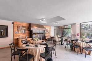 Uma área de refeições com várias mesas e cadeiras, parede de tijolos e cozinha moderna ao fundo.