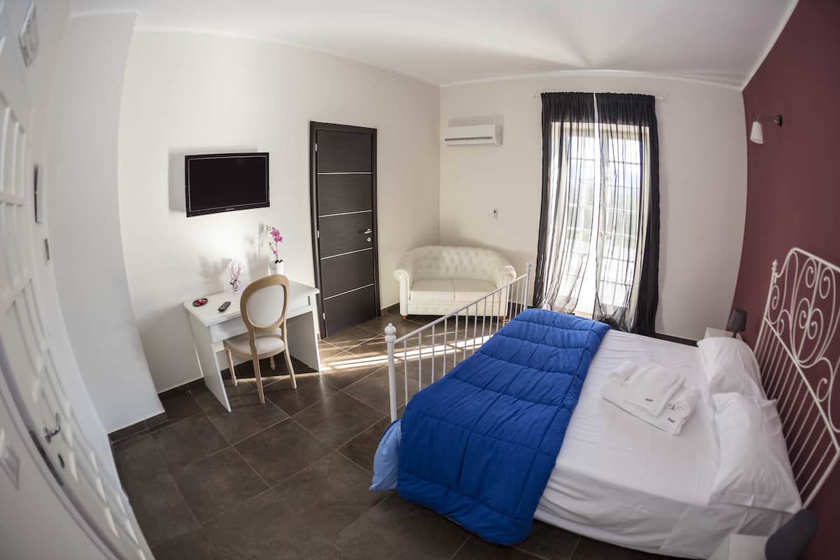 Quarto com cama, escrivaninha, cadeira, televisão e janela com cortinas.
