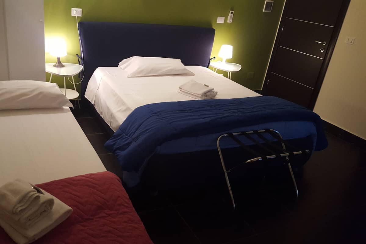 Quarto de hotel com duas camas de solteiro, uma com colcha azul e outra vermelha. Há uma mesinha com abajur entre as camas, e um aquecedor fixado na parede acima delas.