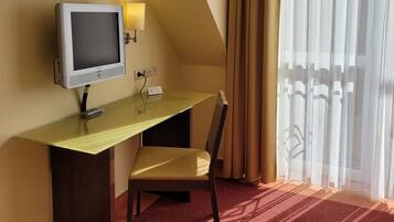 Quarto de hotel com escrivaninha, cadeira, televisão e janela com cortinas.