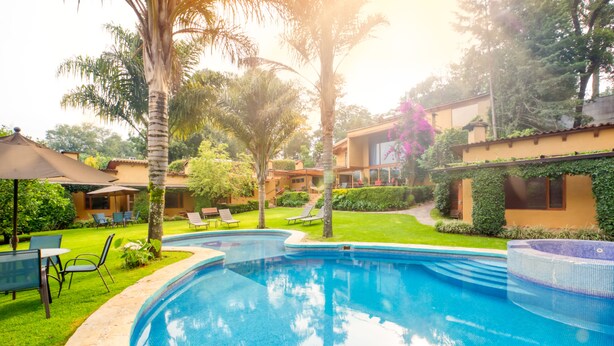 Piscina com espreguiçadeiras e guarda-sóis, cercada por vegetação e edifícios.