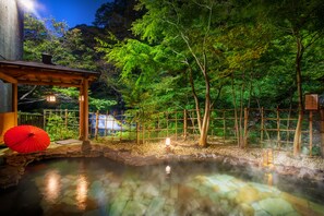 Uma onsen japonesa tradicional com um pavilhão de madeira, uma fonte termal de pedra e um guarda-sol vermelho.