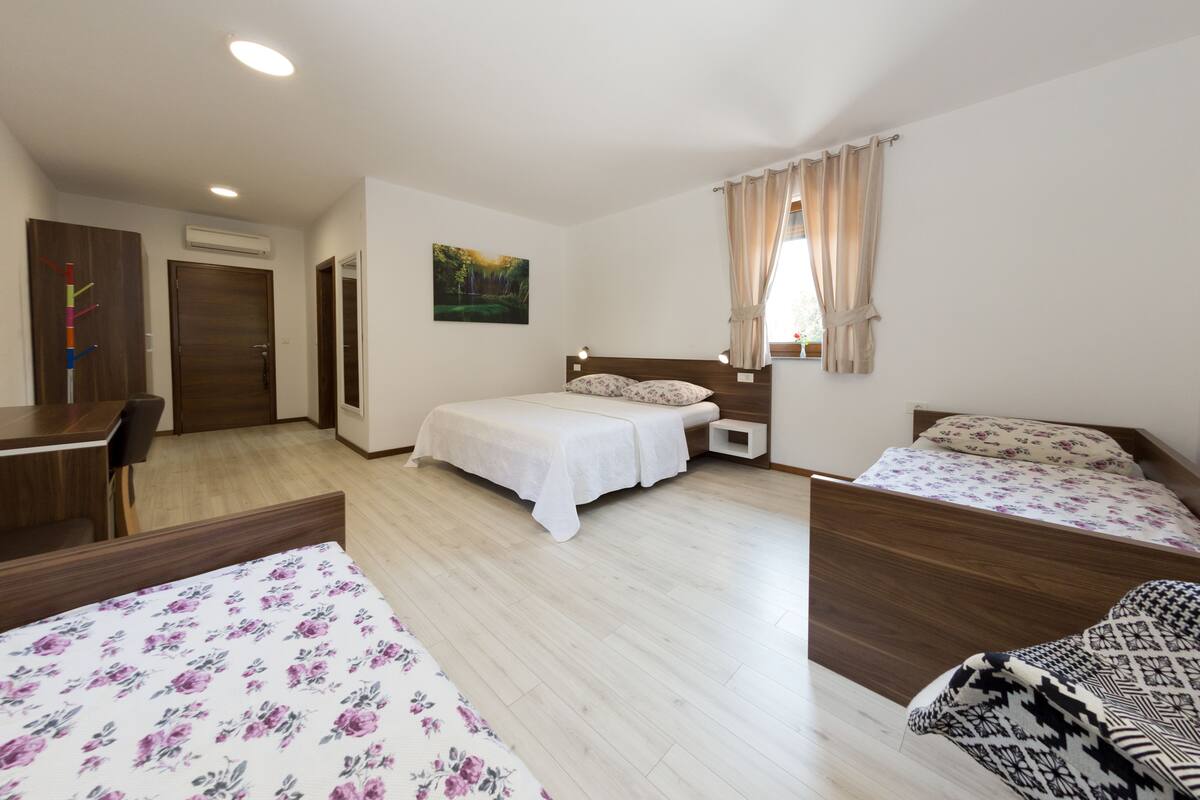 Quarto com duas camas, uma mesa e uma cadeira. Há uma pintura na parede e uma janela com cortinas.