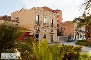 Um edifício de vários andares com varandas, um letreiro com 'La Suite Bed & Breakfast' e uma rua com carros e palmeiras.