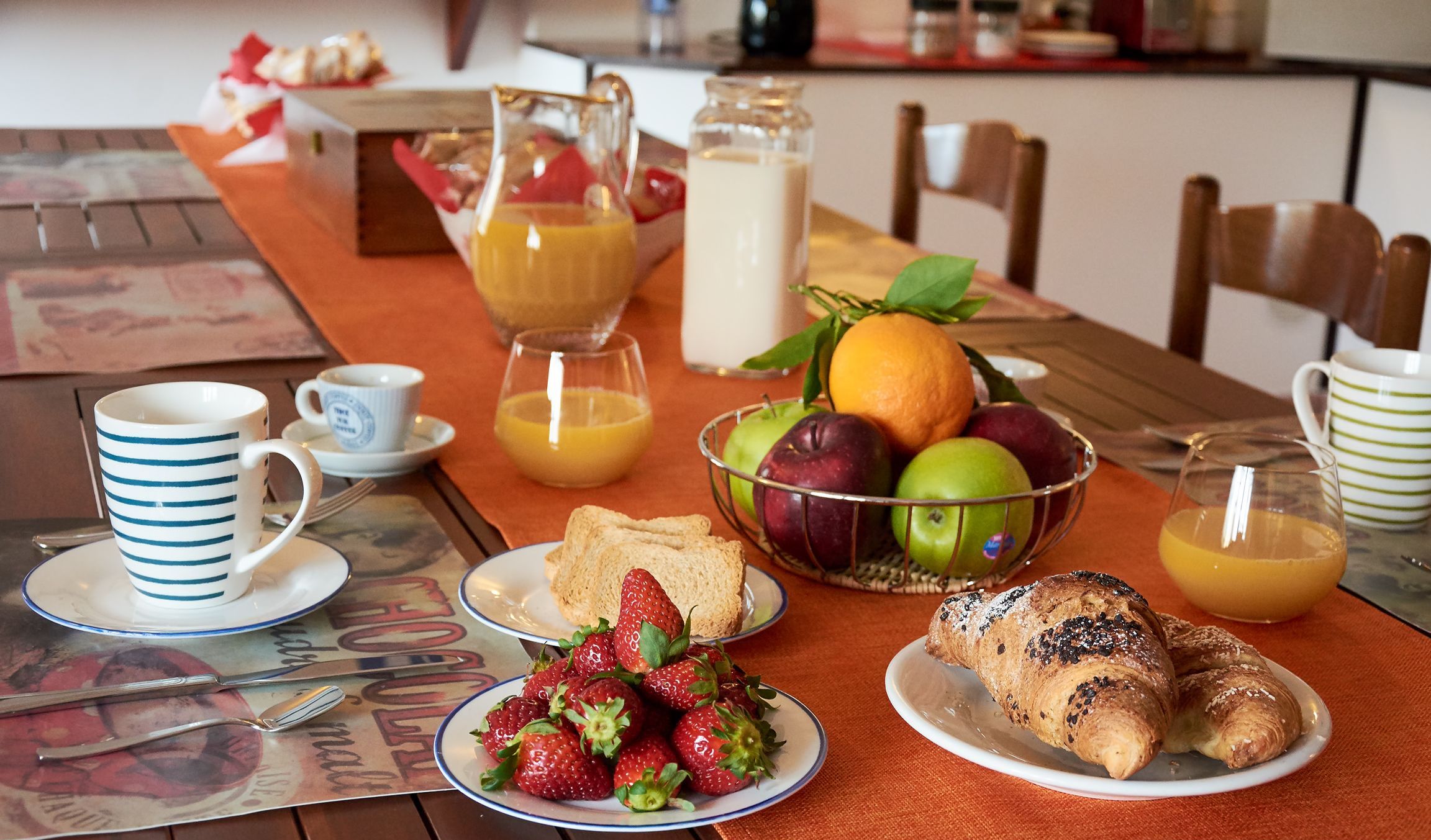 Uma mesa de café da manhã com pão, morangos, um croissant, uma saladeira de frutas e suco de laranja.