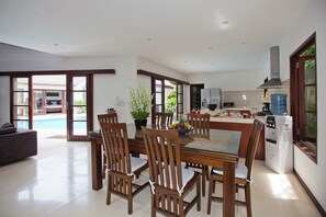 Uma sala de jantar espaçosa com mesa de vidro e cadeiras de madeira, uma cozinha com eletrodomésticos modernos e vista para uma piscina.