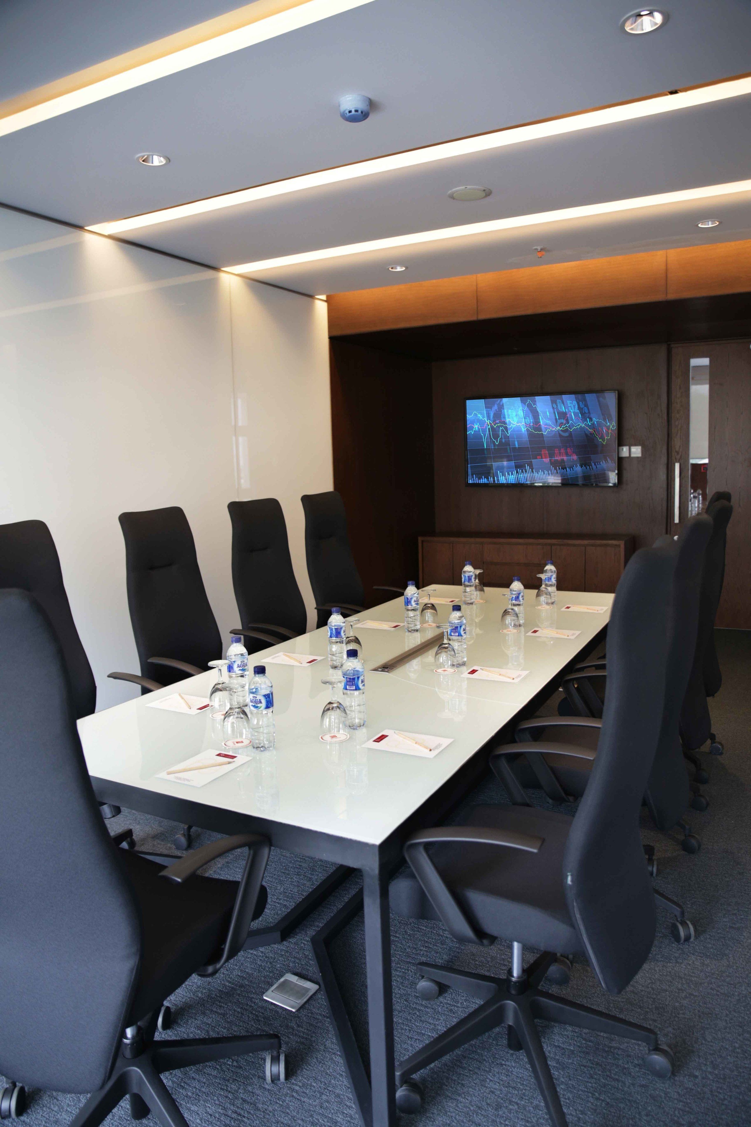 Uma sala de conferências com uma mesa comprida, cadeiras pretas, garrafas de água e uma TV exibindo gráficos financeiros.