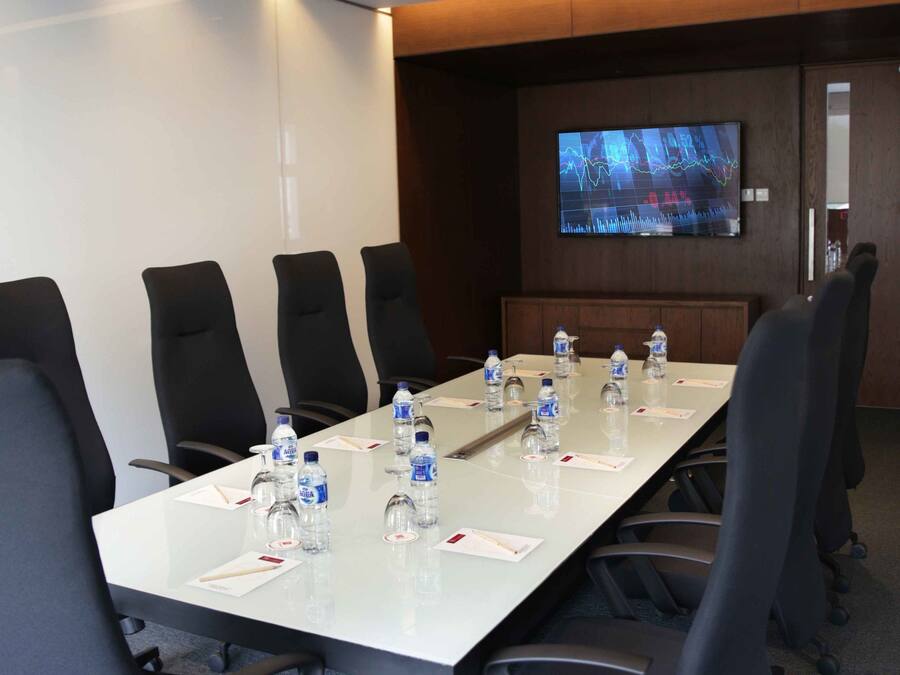 Uma sala de conferências com uma mesa comprida, cadeiras pretas, garrafas de água e uma TV exibindo gráficos financeiros.