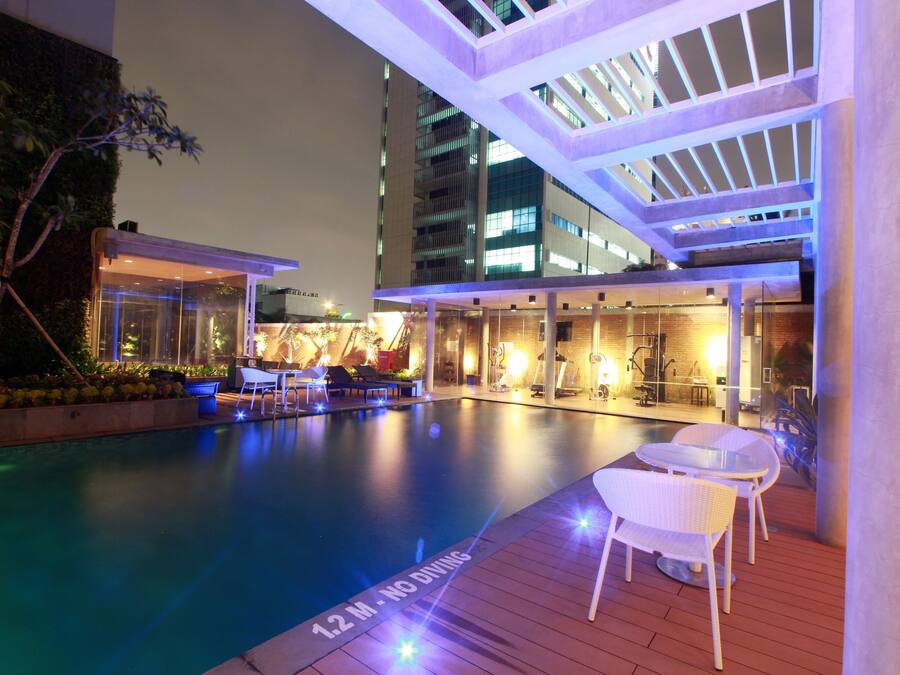 Uma área de piscina à noite, com um compartimento de vidro, assentos externos e um edifício com janelas iluminadas ao fundo.