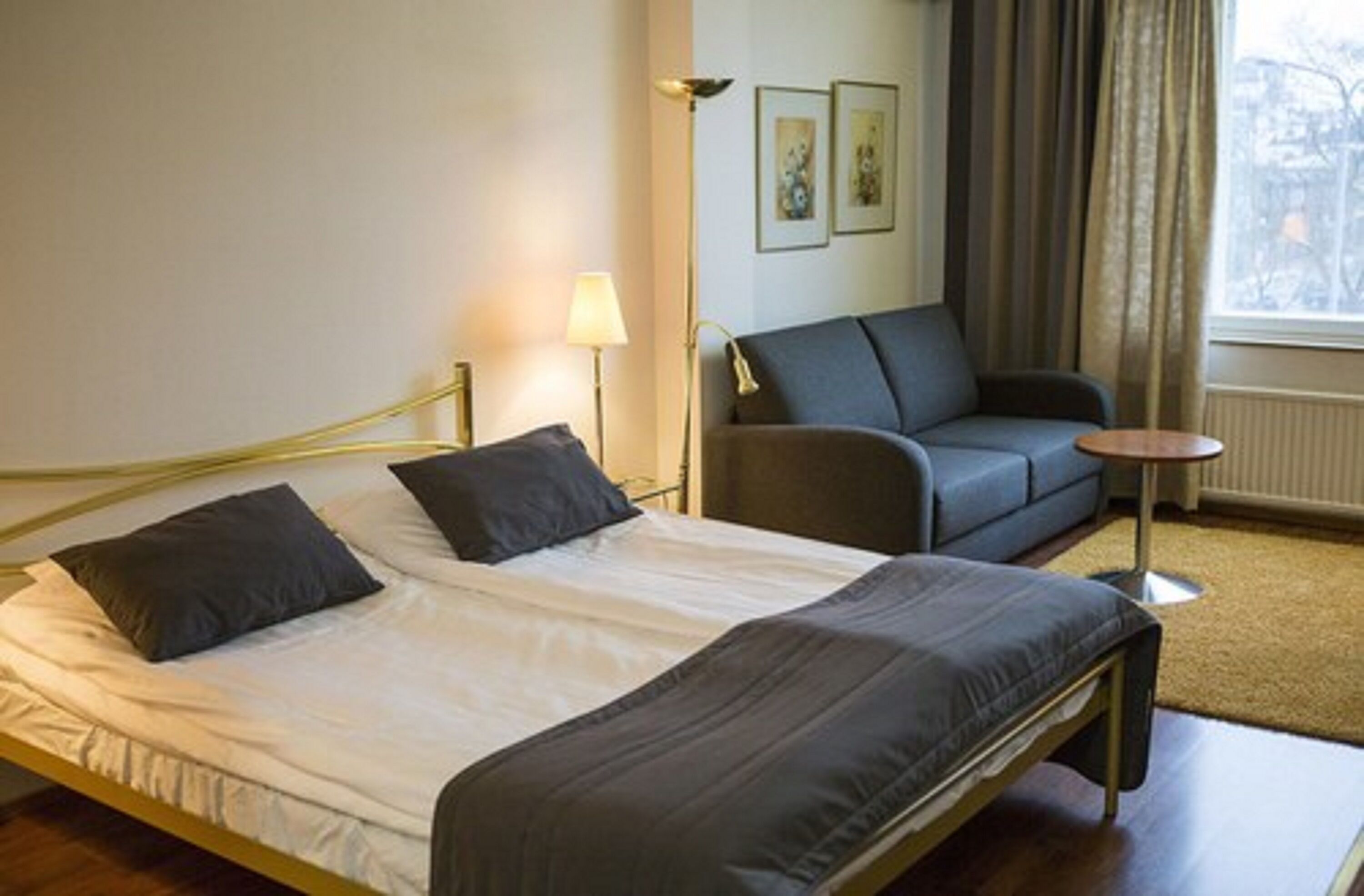 Quarto de hotel com cama, sofá, luminária e janela com cortinas.
