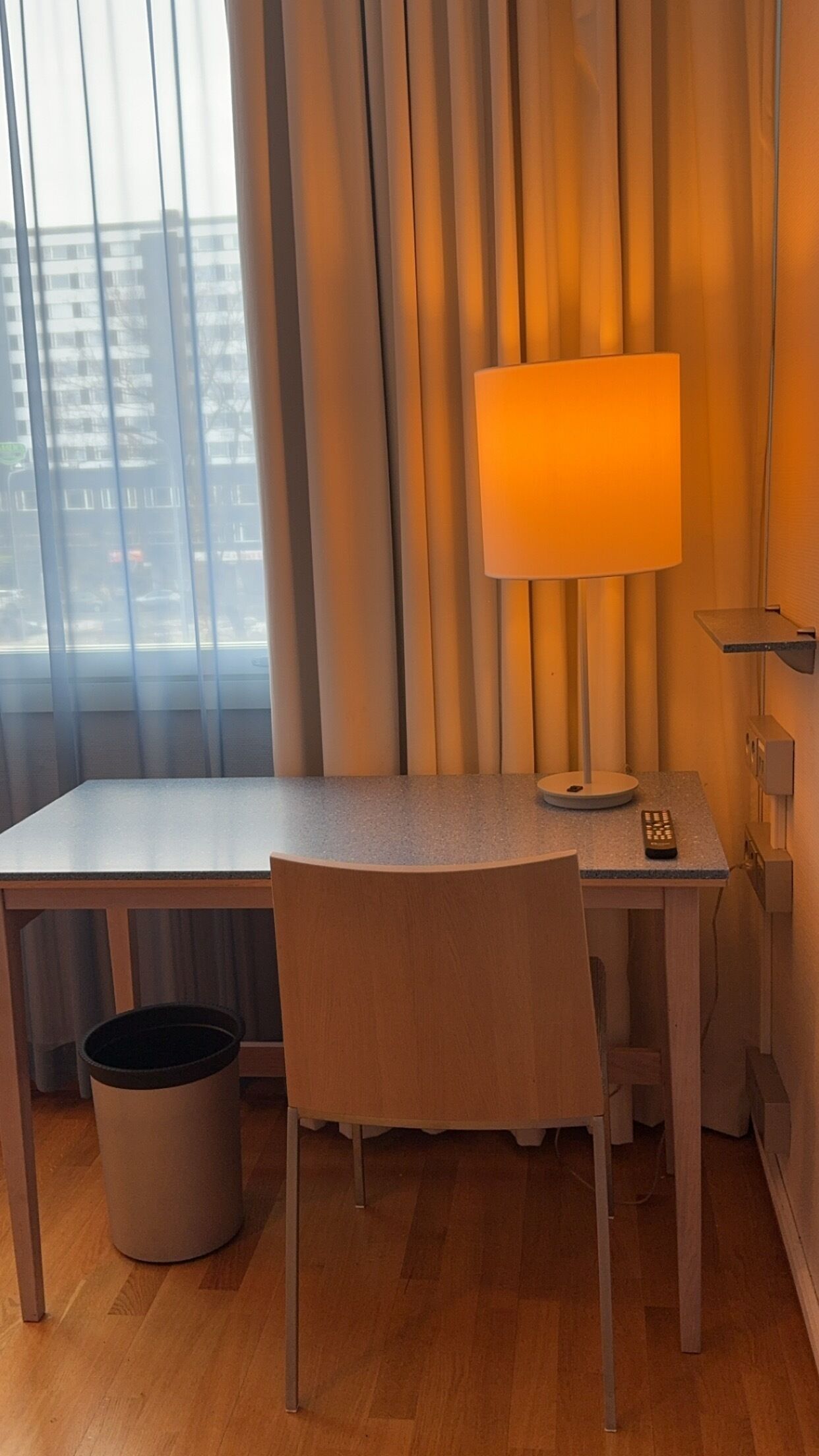 Quarto de hotel com escrivaninha, cadeira, luminária e lixeira.