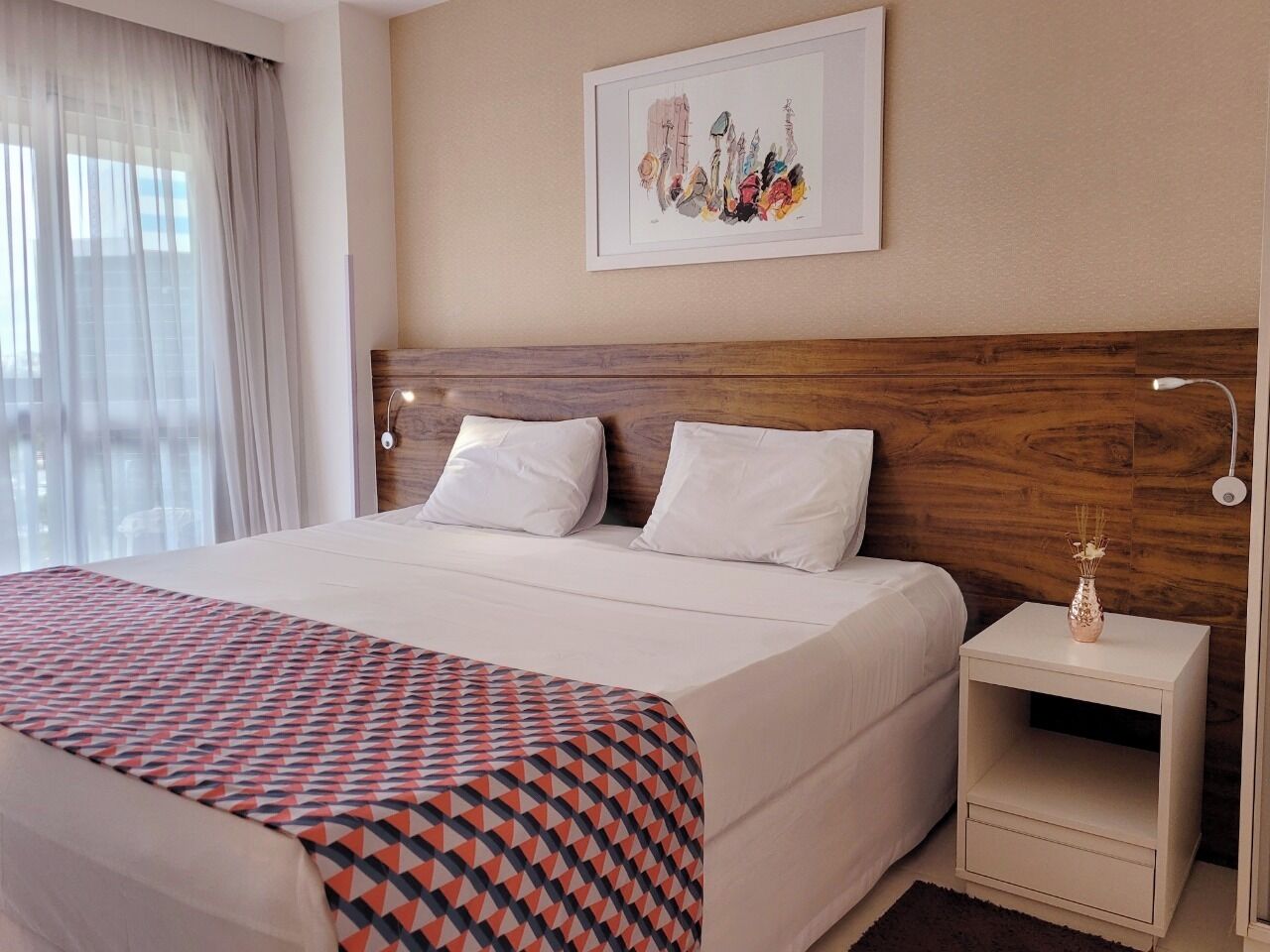 Quarto de hotel com uma cama grande, cabeceira de madeira, mesa de cabeceira com um vaso e uma moldura na parede.